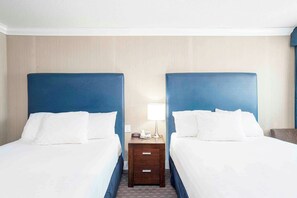 Quarto de hotel com duas camas, uma mesa de cabeceira com abajur e um móvel de gavetas de madeira.