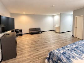 Quarto de hotel moderno com televisão de tela plana, sofá, pequeno espaço de cozinha e cama com colcha estampada.