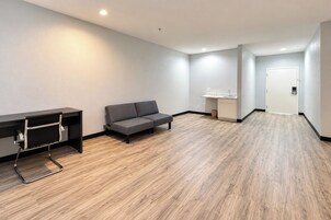 Quarto moderno e minimalista com piso de madeira, sofá cinza, escrivaninha com cadeira e pequena área de cozinha.