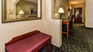 Corredor de hotel com um banco, uma mesa com abajur, um espelho e uma porta.