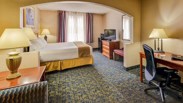 Quarto de hotel com cama, escrivaninha, cadeira, televisão e luminárias.