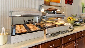 Uma área de buffet com diversos alimentos, incluindo waffles e doces, e uma saladeira de frutas.