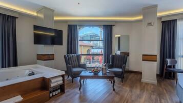 Quarto de hotel moderno com uma janela ampla, televisão de tela plana, sofá, duas poltronas, mesa de centro e minibar.