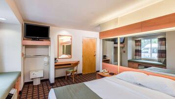 Quarto de hotel com cama, escrivaninha com cadeira, televisão, espelho, janela com cortinas e uma pequena mesa com telefone e abajur.