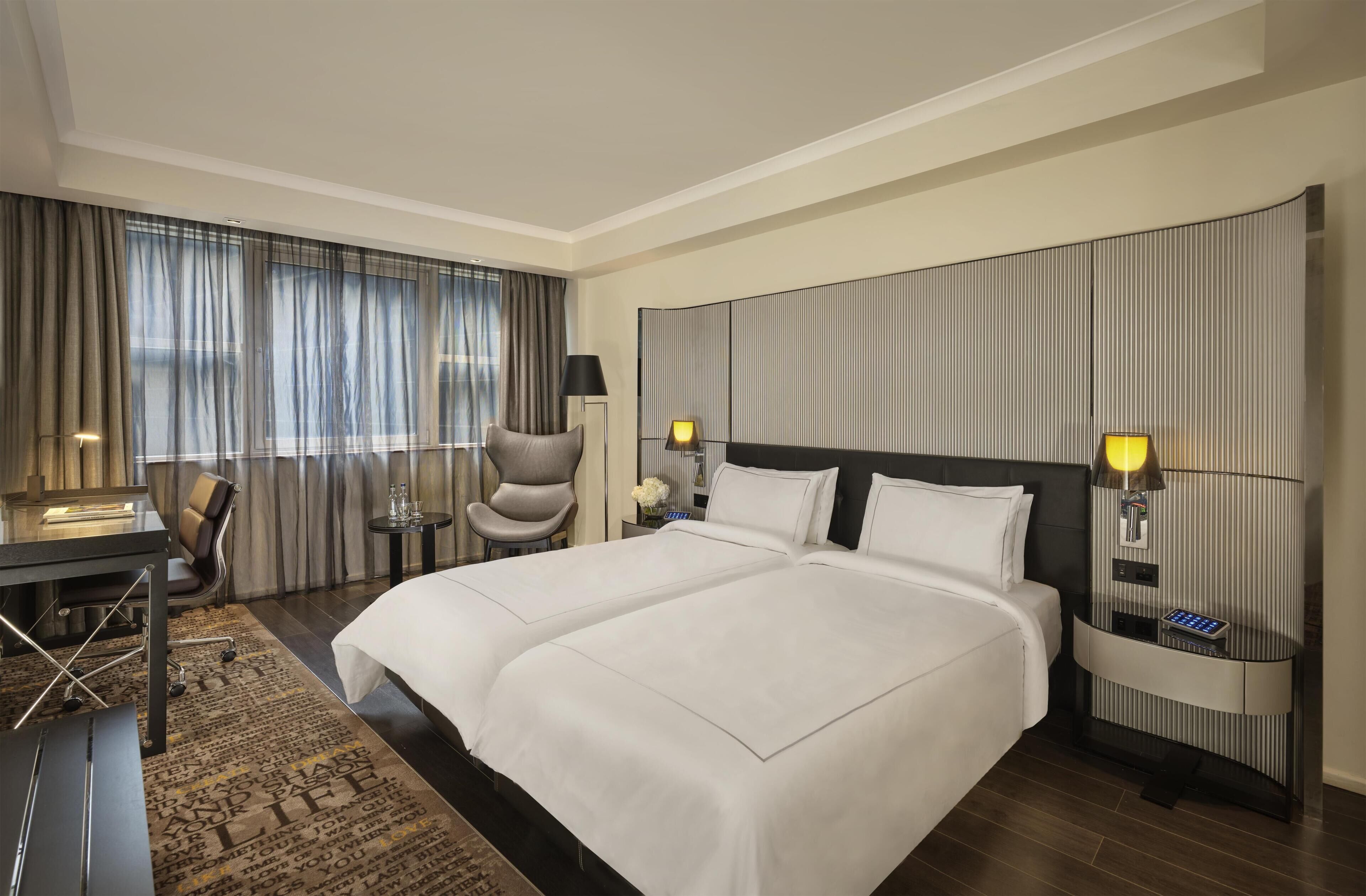 Quarto de hotel com uma cama grande, uma escrivaninha, uma cadeira, uma mesinha de cabeceira e uma luminária.