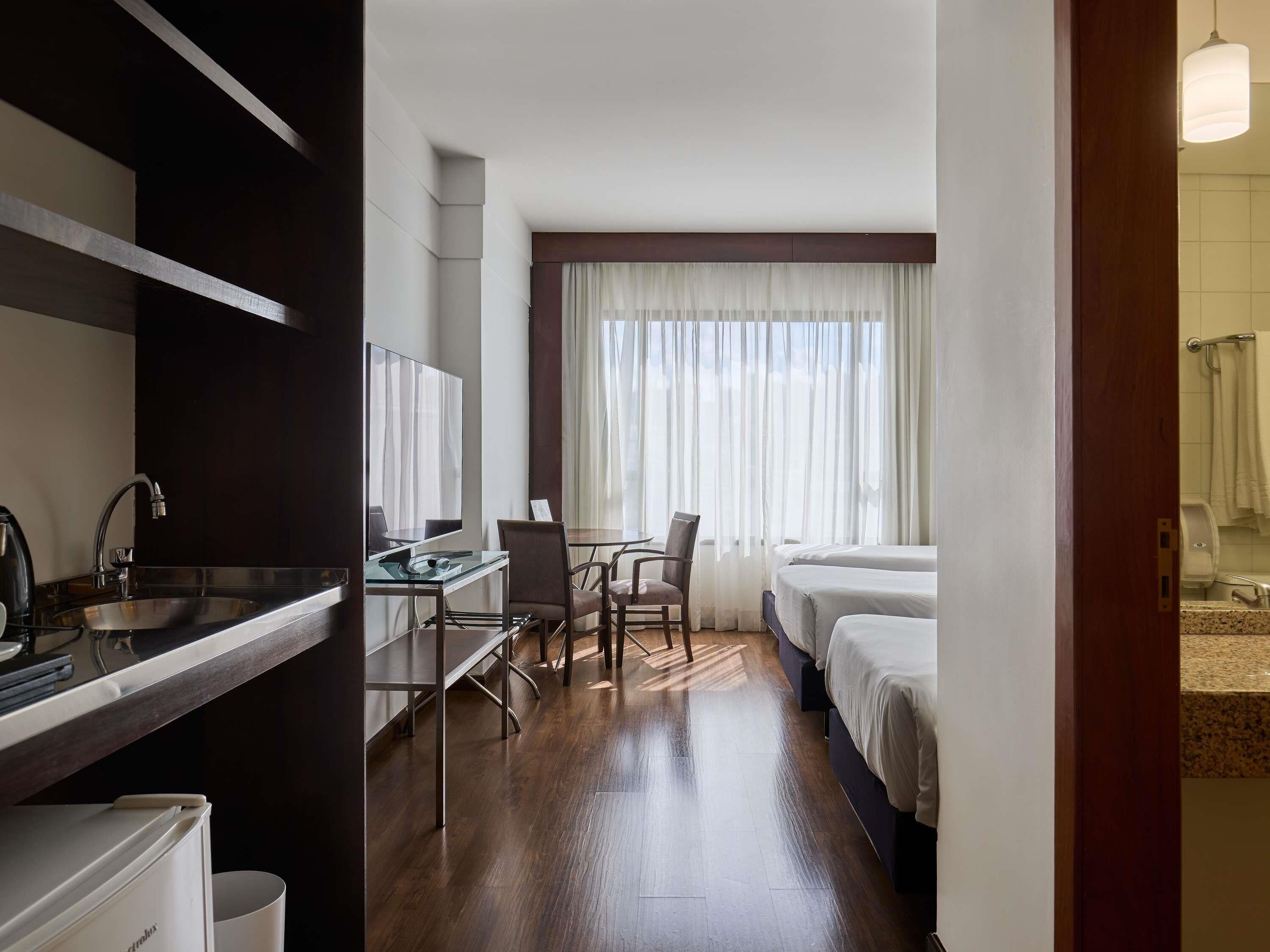 Quarto de hotel com escrivaninha, duas camas, janela com cortinas e cozinha compacta.