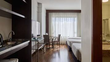 Quarto de hotel com escrivaninha, duas camas, janela com cortinas e cozinha compacta.
