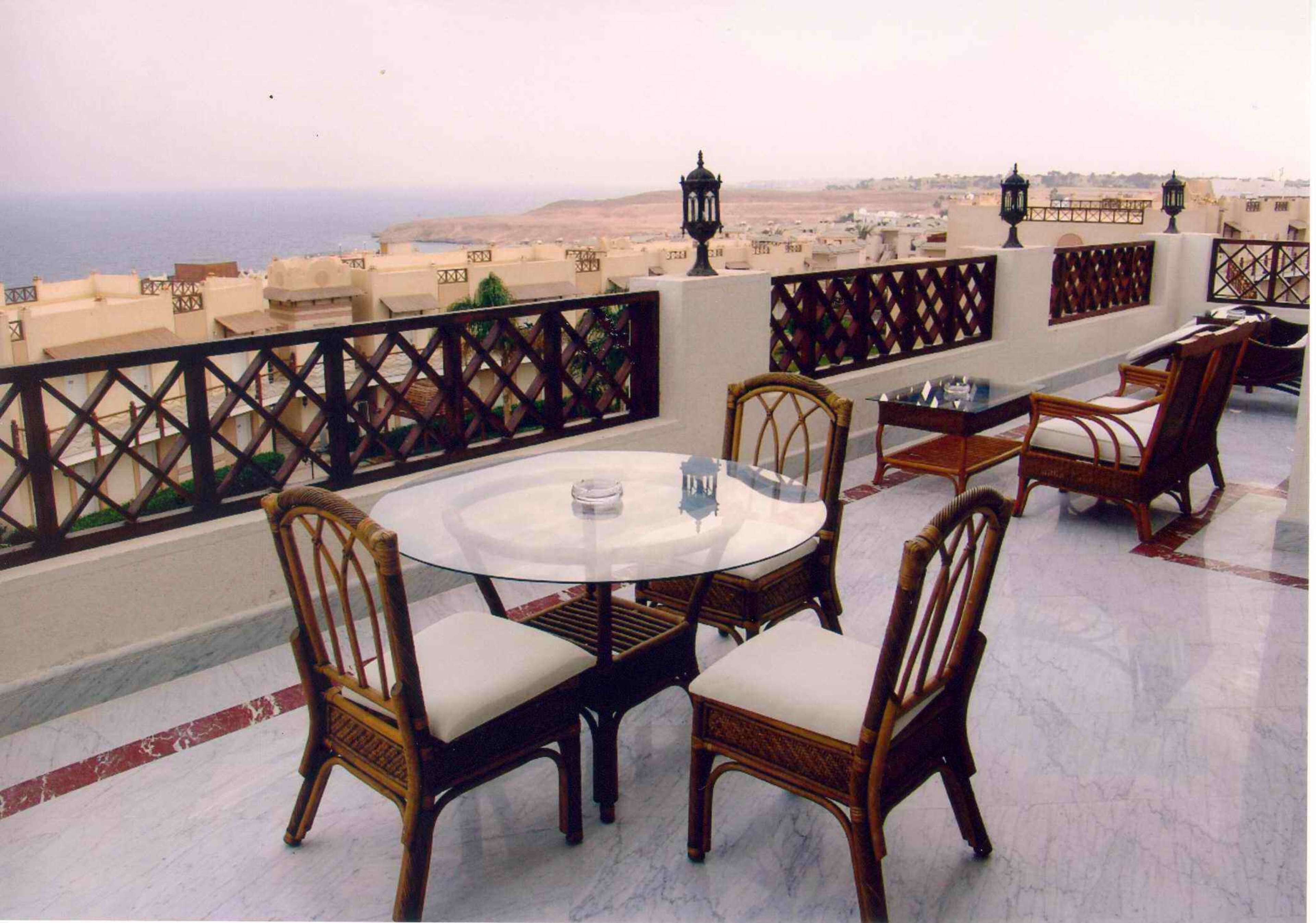 Uma mesa redonda com quatro cadeiras de vime e um tampo de vidro, disposta num terraço com vista para a cidade e para o mar.