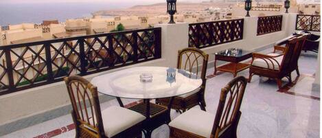Uma mesa redonda com quatro cadeiras de vime e um tampo de vidro, disposta num terraço com vista para a cidade e para o mar.