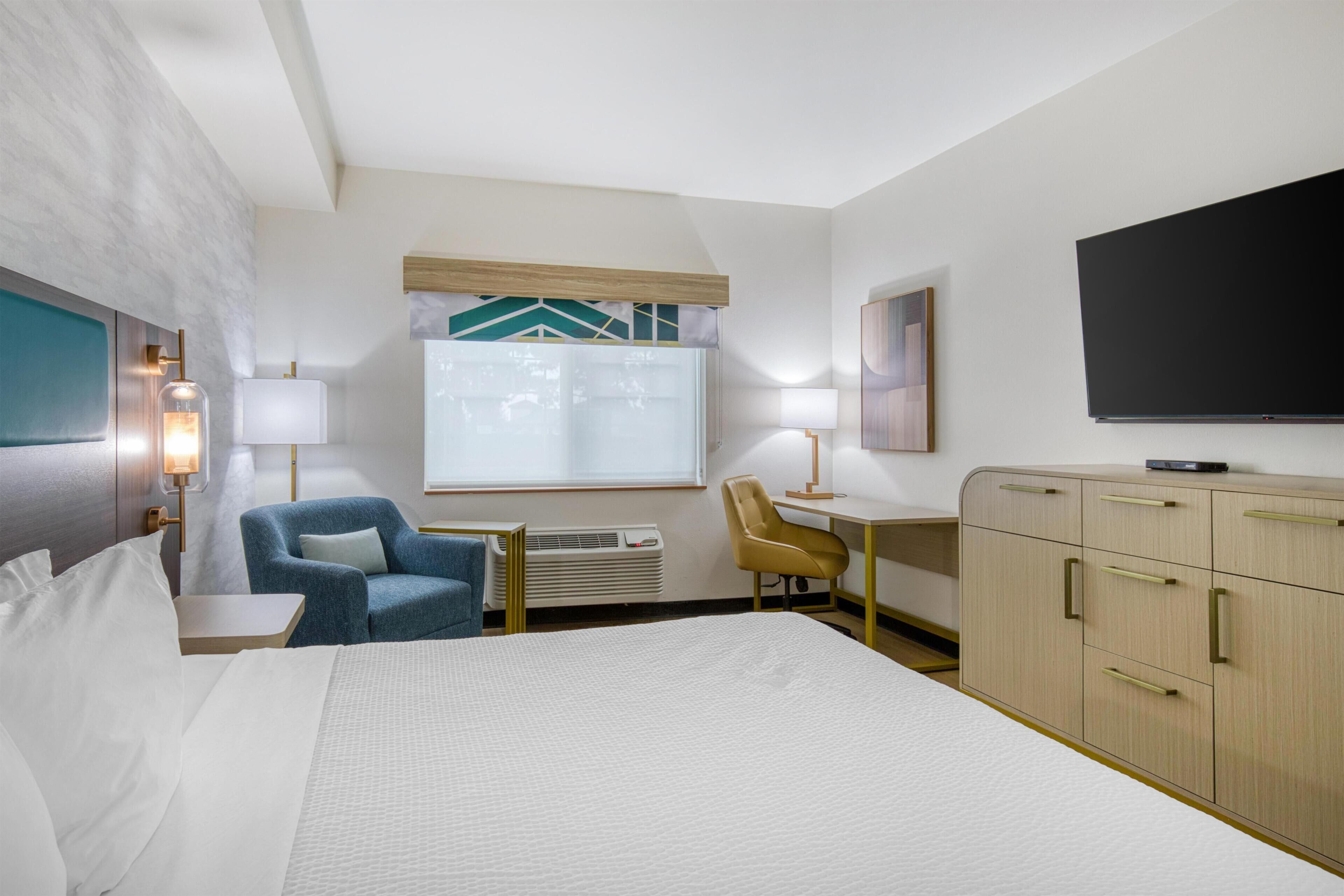 Quarto de hotel com uma cama, uma escrivaninha, uma cadeira, uma televisão e um guarda-roupa.