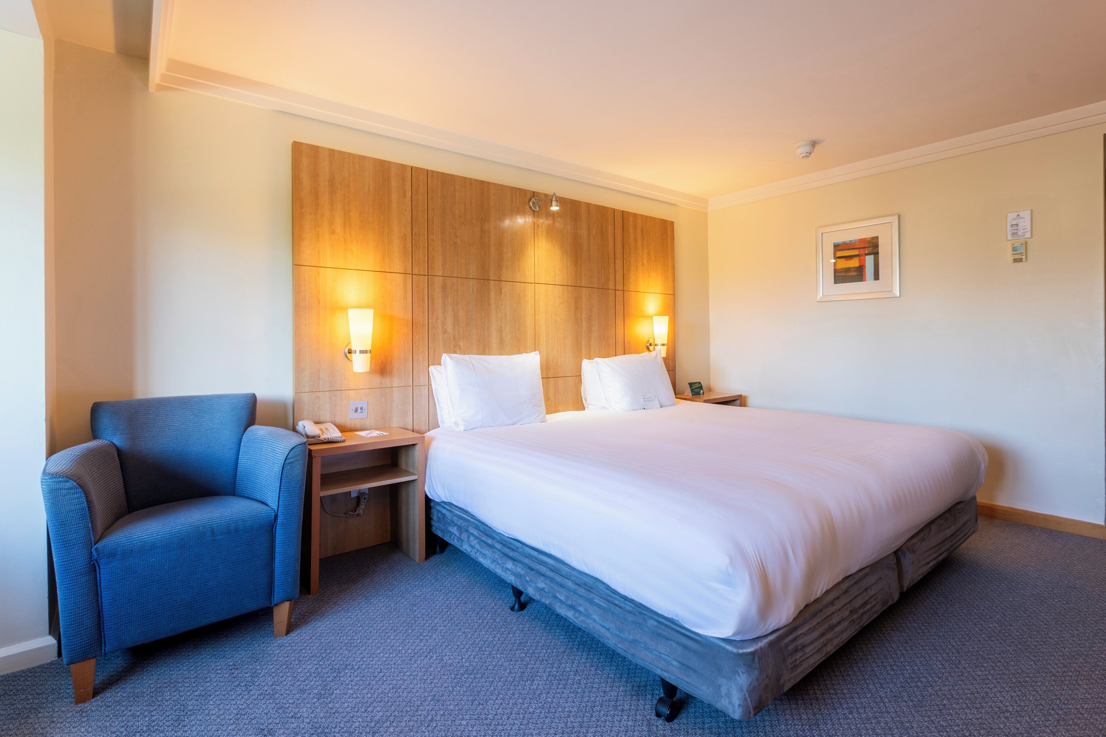 Quarto de hotel com uma cama grande, uma cadeira, mesa de cabeceira com telefone e duas luminárias fixadas na parede.