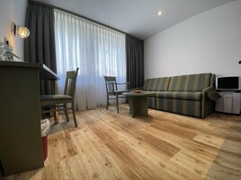 Uma sala de estar moderna com piso de madeira, um sofá, duas poltronas, uma mesa pequena e um espaço com cozinha compacta.