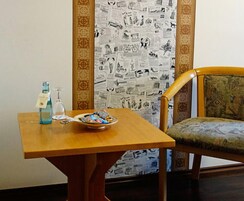 Uma mesa de madeira com uma garrafa e uma tigela de petiscos, uma cadeira com almofada estampada, e uma parede com padrão de jornal.