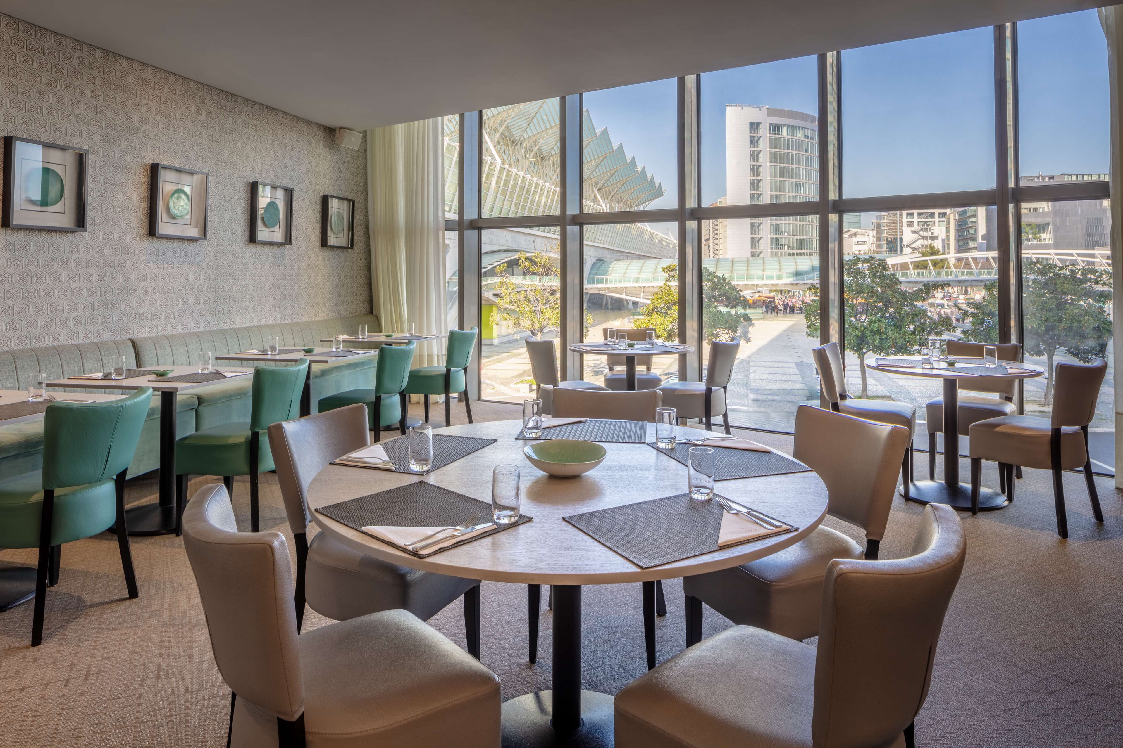 Uma sala de jantar moderna com mesas e cadeiras redondas, obra de arte emoldurada na parede e vista para a cidade através de grandes janelas.