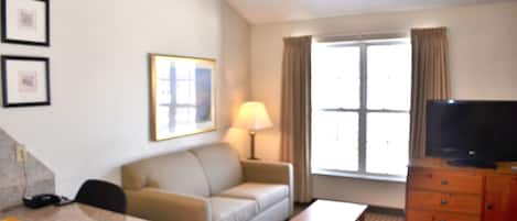 Quarto de hotel com sofá, mesa pequena, televisão e janela com cortinas.