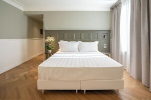 Uma cama bem arrumada em quarto de hotel, com piso de madeira, cabeceira cinza e mesa de cabeceira com um vaso de flores.