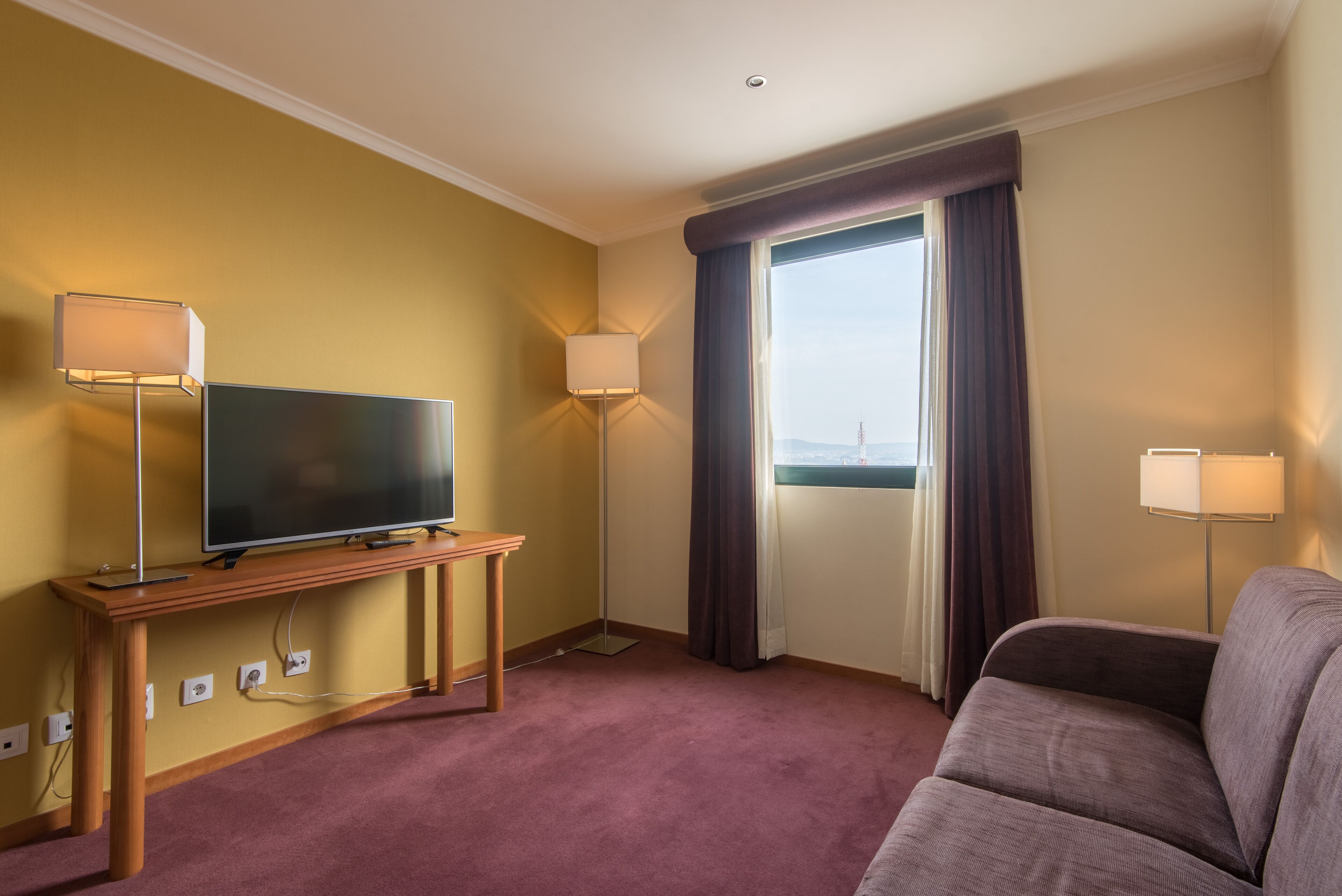 Quarto de hotel com televisão sobre suporte de madeira, sofá e uma janela grande com cortinas.