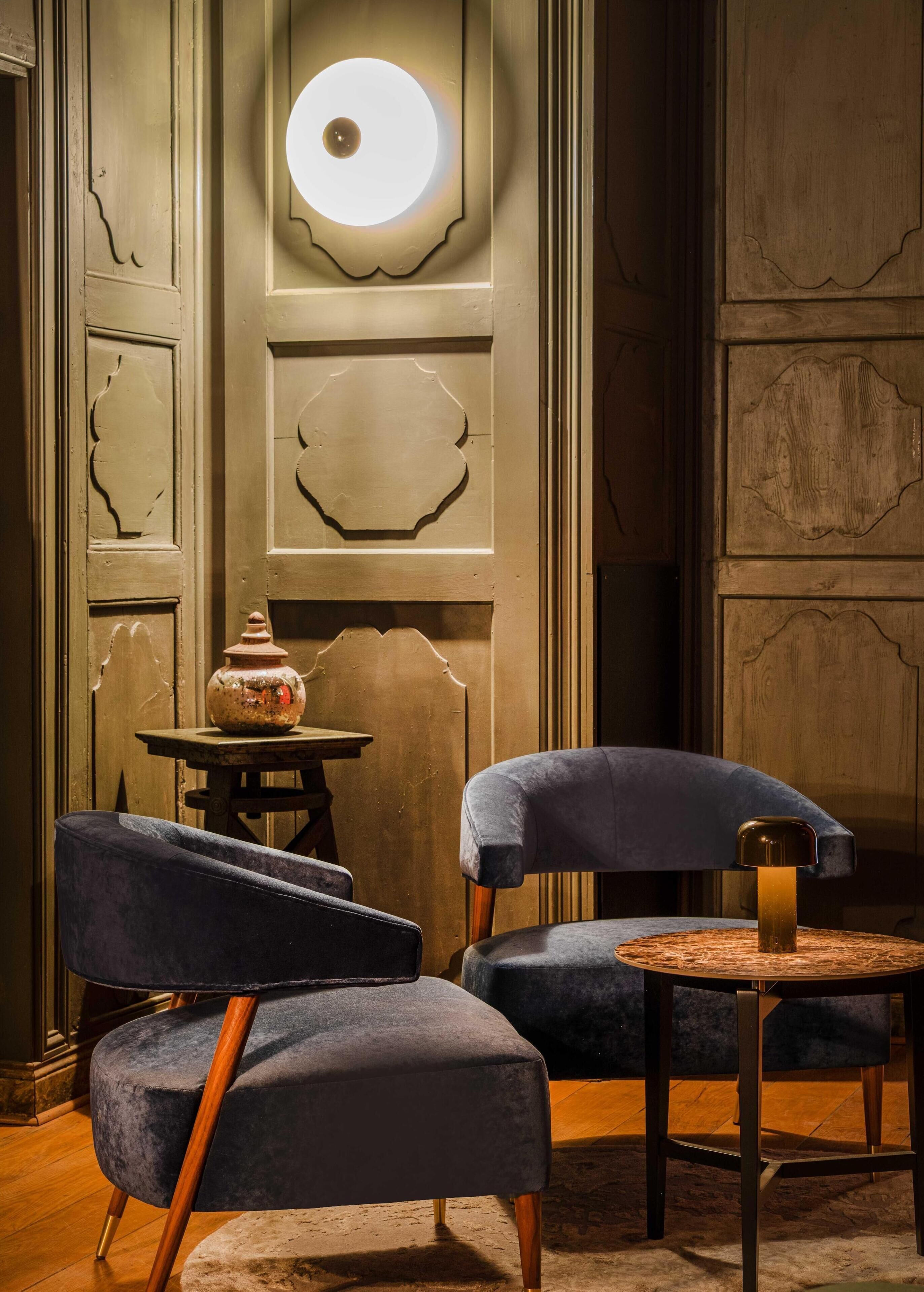 Um quarto com duas poltronas estofadas em azul escuro, uma mesa redonda pequena com um vaso de cerâmica, e uma parede com painéis decorativos e um candeeiro circular.