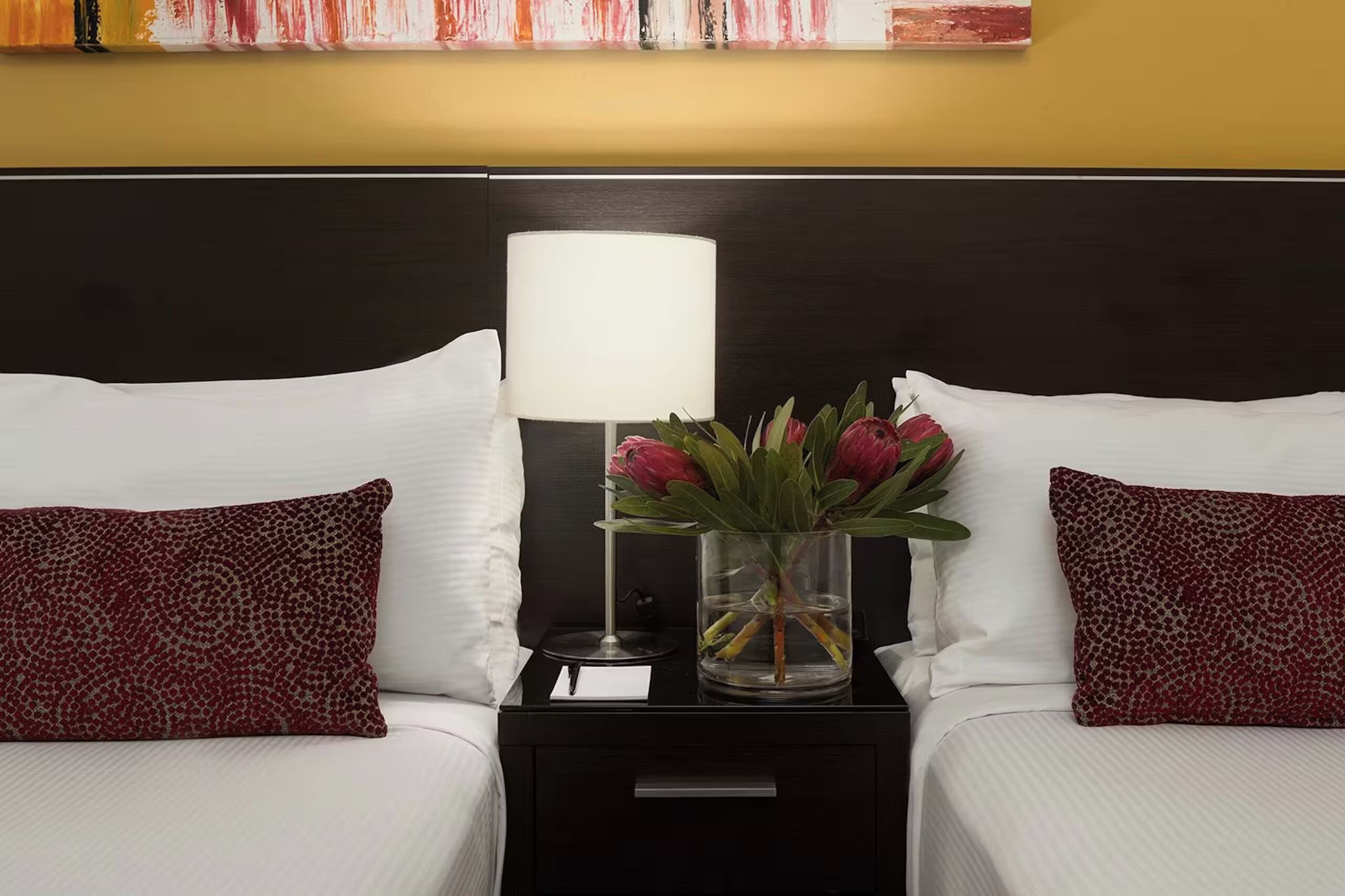 Quarto de hotel com duas camas, uma mesa de cabeceira com abajur e um vaso com flores.