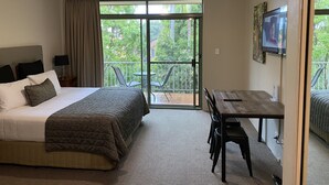Quarto de hotel com cama, escrivaninha, televisão e varanda com vista para árvores.
