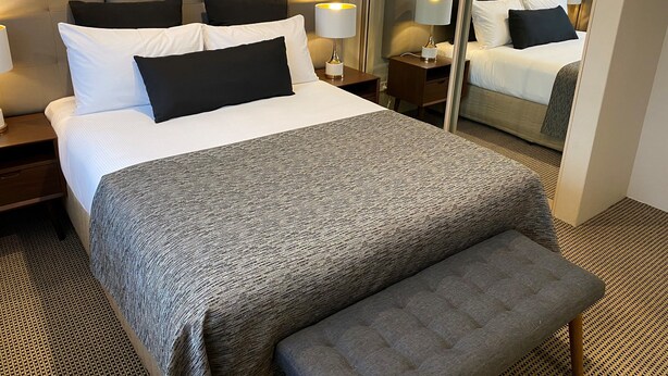 Quarto de hotel com uma cama grande, uma mesinha de cabeceira com abajur, um espelho e um banco.