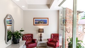 Um hall com duas poltronas de couro vermelho, uma mesa branca com abajur, um quadro na parede e um espelho à esquerda.