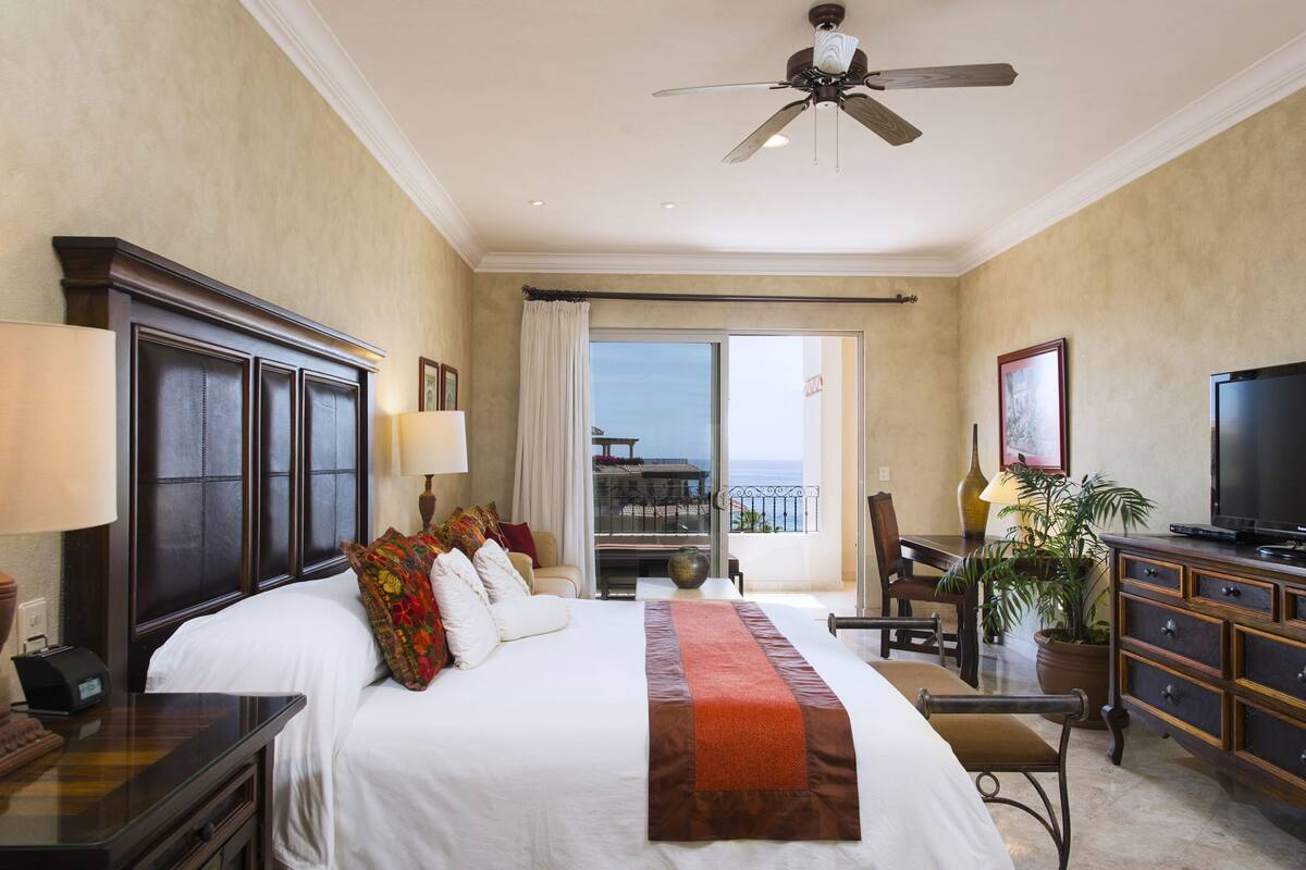 Quarto de hotel com cama, escrivaninha, televisão, ventoinha no teto e varanda com vista.