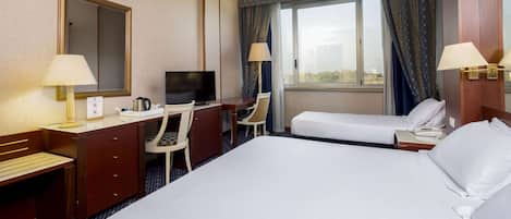 Quarto de hotel com uma cama grande, uma escrivaninha com televisão, uma cadeira, abajur e janela com cortinas.