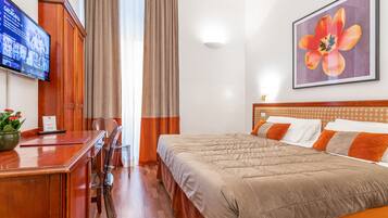 Quarto de hotel com uma cama grande, uma escrivaninha com computador, uma TV e uma pintura de flores na parede.