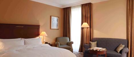 Quarto de hotel com uma cama grande, duas poltronas, uma mesinha e uma janela com cortinas.