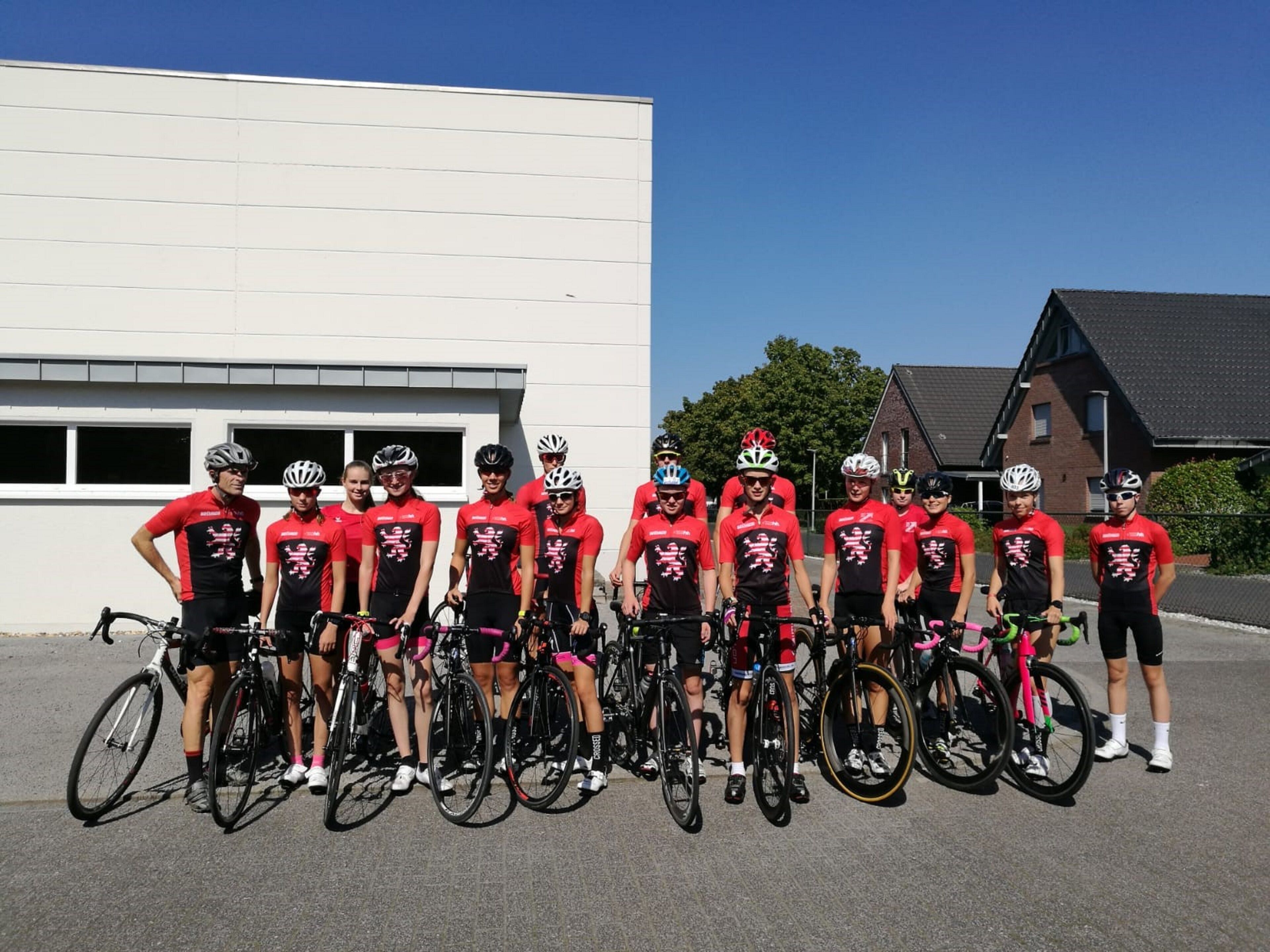 Um grupo de ciclistas em uniformes vermelhos e pretos posa em frente a um edifício branco, com uma casa de tijolos ao fundo.