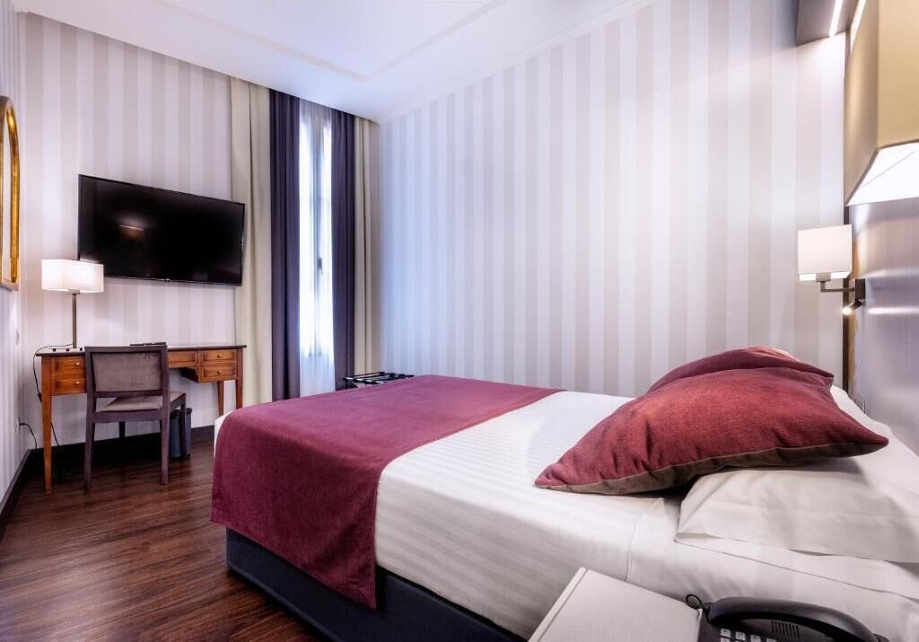 Quarto de hotel com uma cama, uma escrivaninha com cadeira, uma televisão na parede e um telefone.