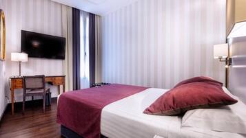 Quarto de hotel com uma cama, uma escrivaninha com cadeira, uma televisão na parede e um telefone.