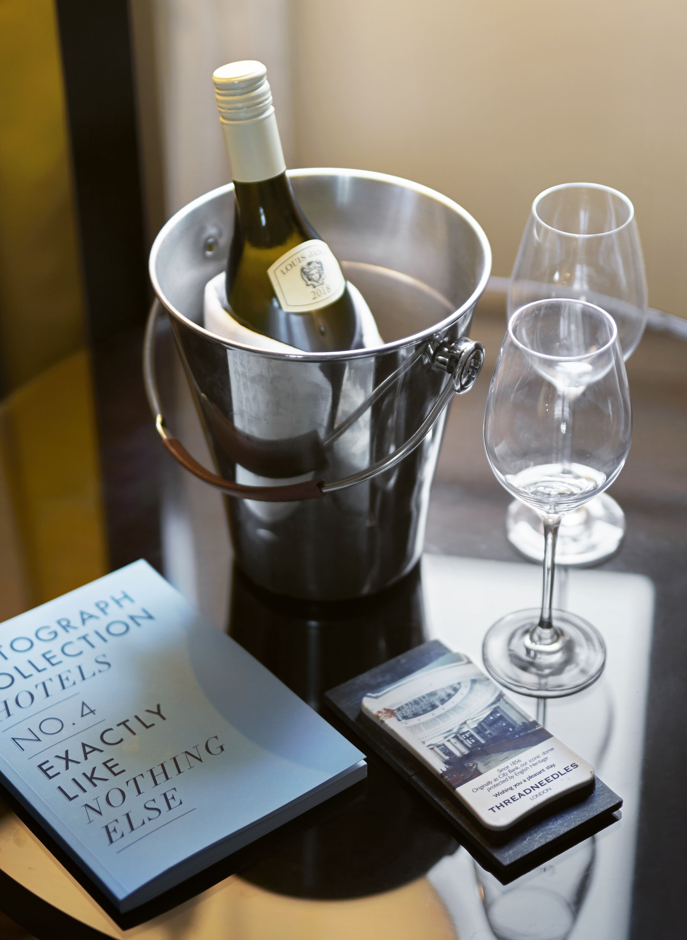 Uma garrafa de vinho em um balde de metal, duas taças de vinho vazias e um livreto intitulado "Exact Like Nothing Else".