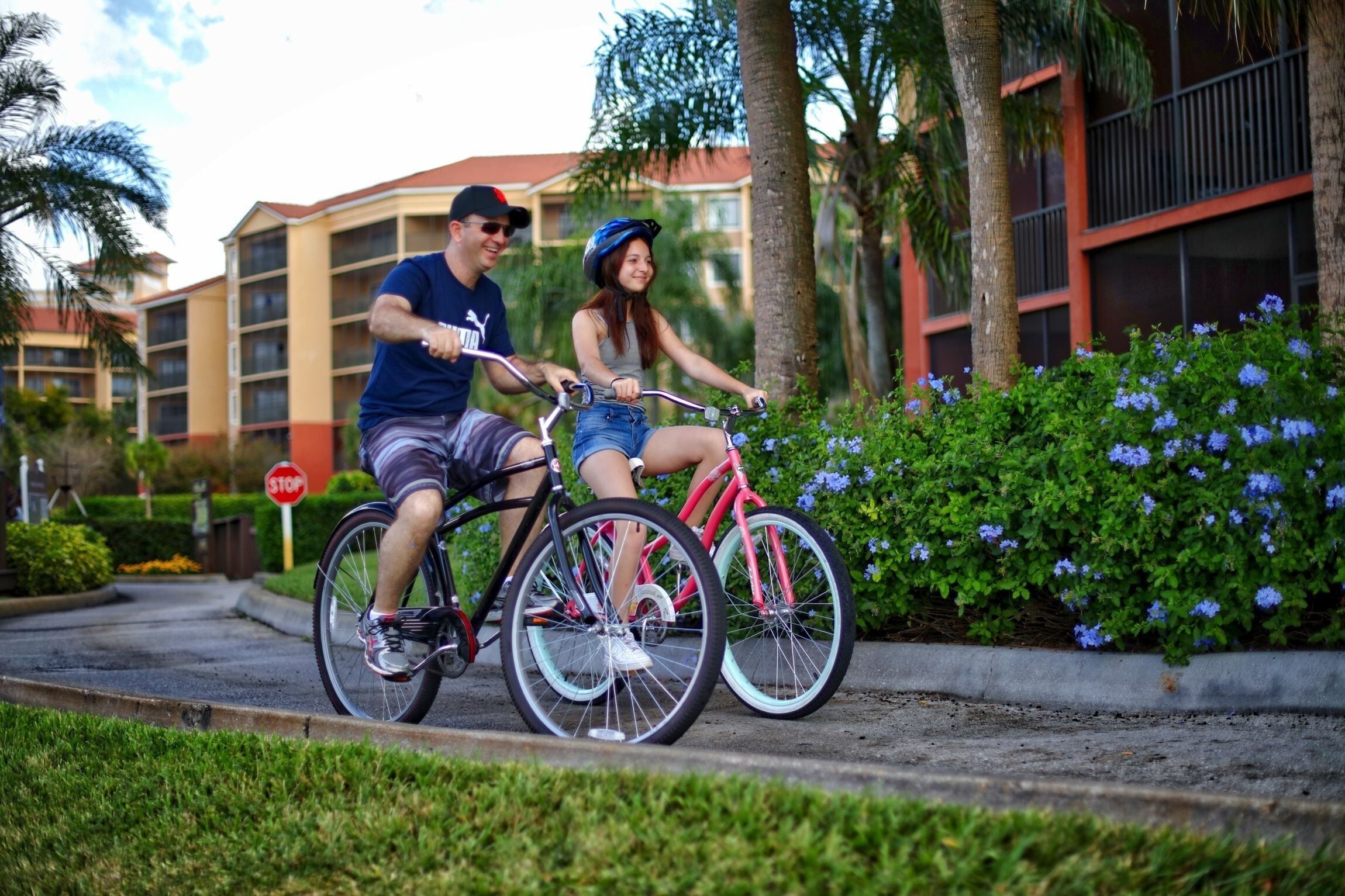 Duas pessoas de bicicleta numa calçada, com uma placa de pare e um edifício ao fundo.