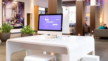 Um lobby de hotel moderno com uma recepção branca, um monitor grande exibindo um site, plantas em vasos e uma área de estar ao fundo.