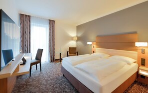 Quarto de hotel com uma cama, uma escrivaninha com cadeira, uma televisão e um abajur.