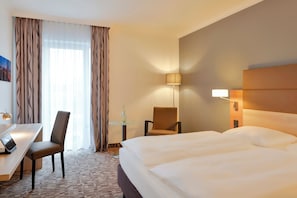Quarto de hotel com cama, escrivaninha com cadeira, abajur e vista para o exterior pela janela.