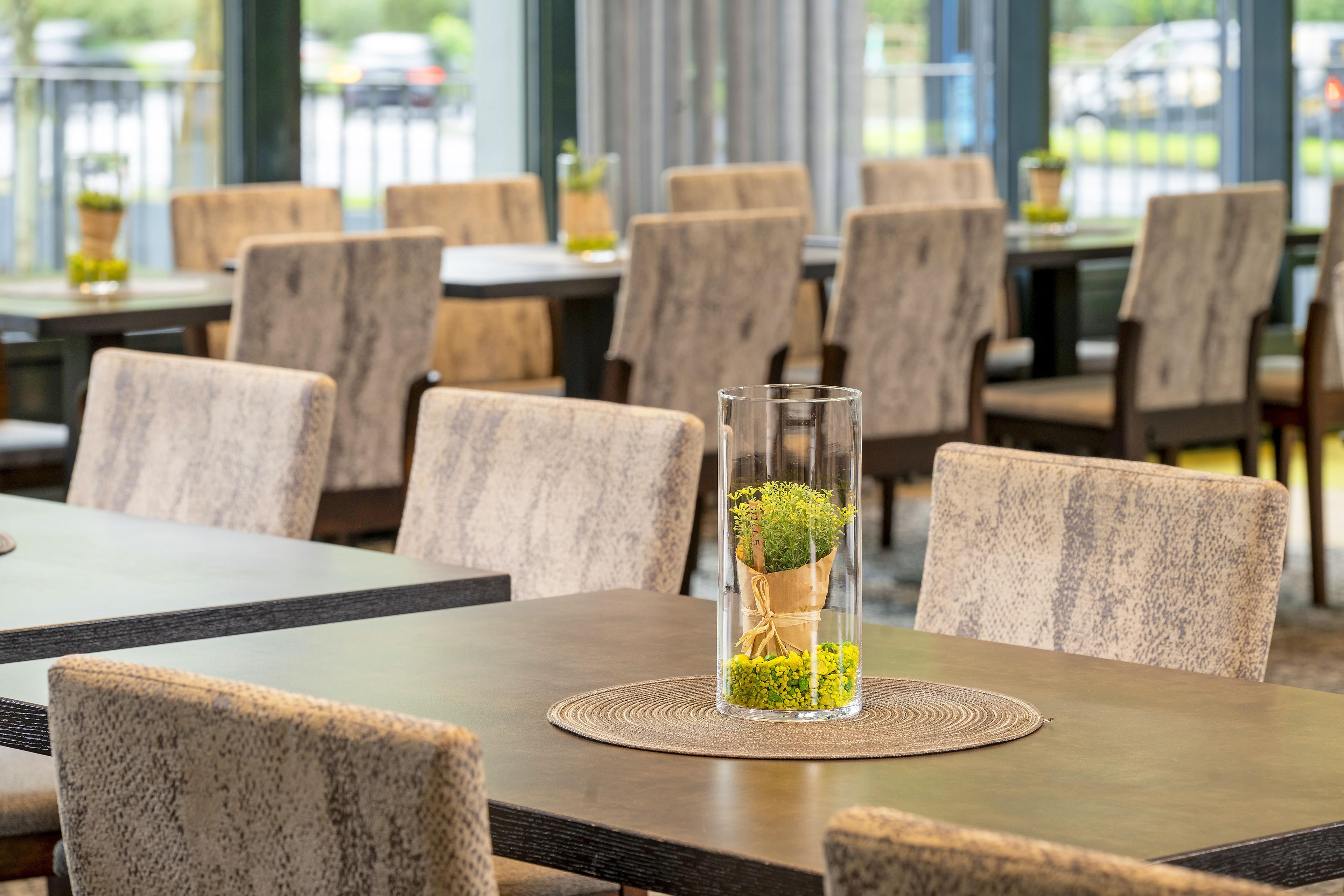 Interior de restaurante com mesas e cadeiras de madeira, vaso de vidro com planta em um jogo americano de palha.
