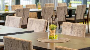 Interior de restaurante com mesas e cadeiras de madeira, vaso de vidro com planta em um jogo americano de palha.