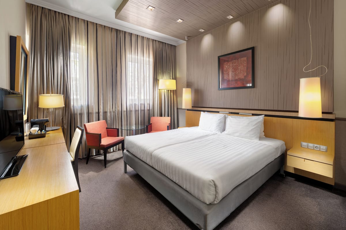 Quarto de hotel com cama, escrivaninha, cadeira, televisão e uma janela grande com cortinas.