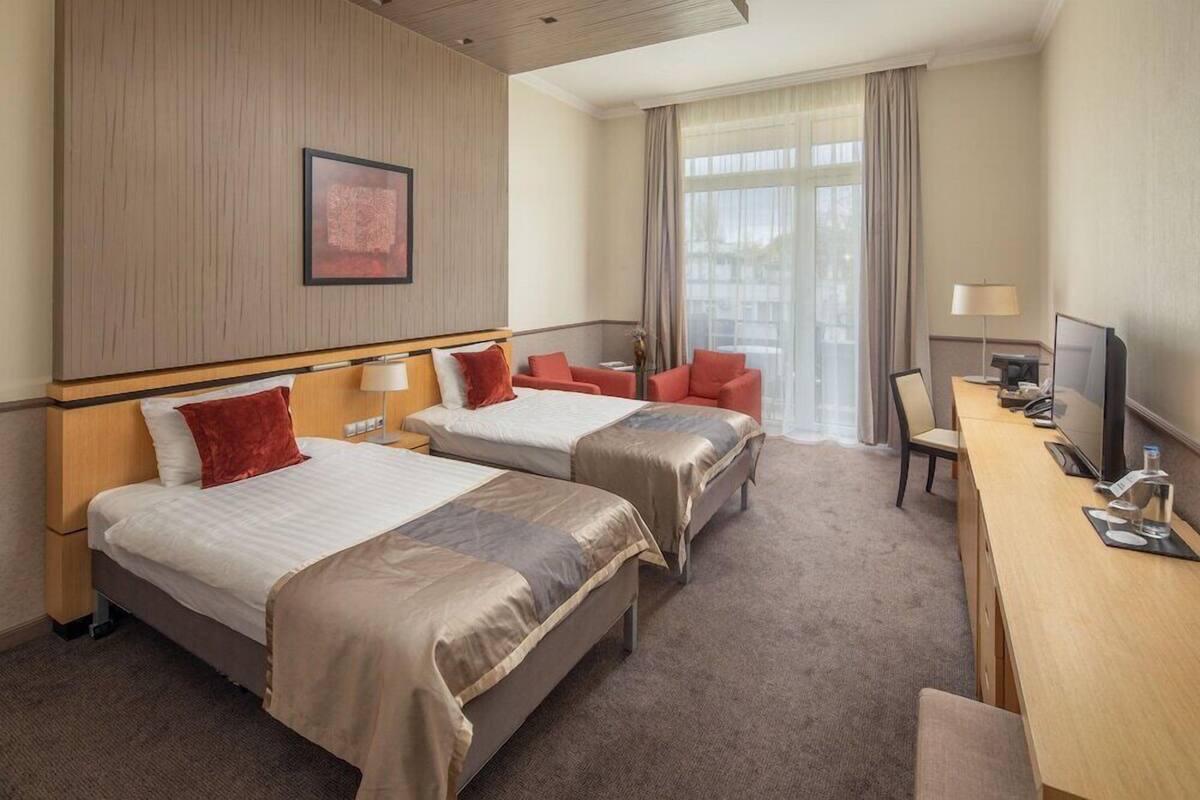 Quarto de hotel com duas camas, uma escrivaninha, uma cadeira, uma televisão e uma janela com cortinas.