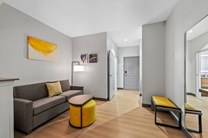 Quarto de hotel moderno com um sofá cinzento, um banquinho amarelo, uma mesinha e um espelho grande.