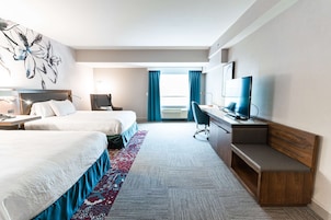 Quarto de hotel com duas camas, uma escrivaninha, uma cadeira, uma televisão e uma janela grande com cortinas.