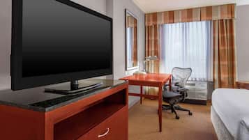 Quarto de hotel com televisão de tela plana, escrivaninha de madeira, cadeira, cama e janela com cortinas.