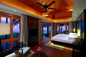 Quarto de hotel com cama, televisão, ventoinha no teto e janelas amplas com vista para palmeiras e o oceano.
