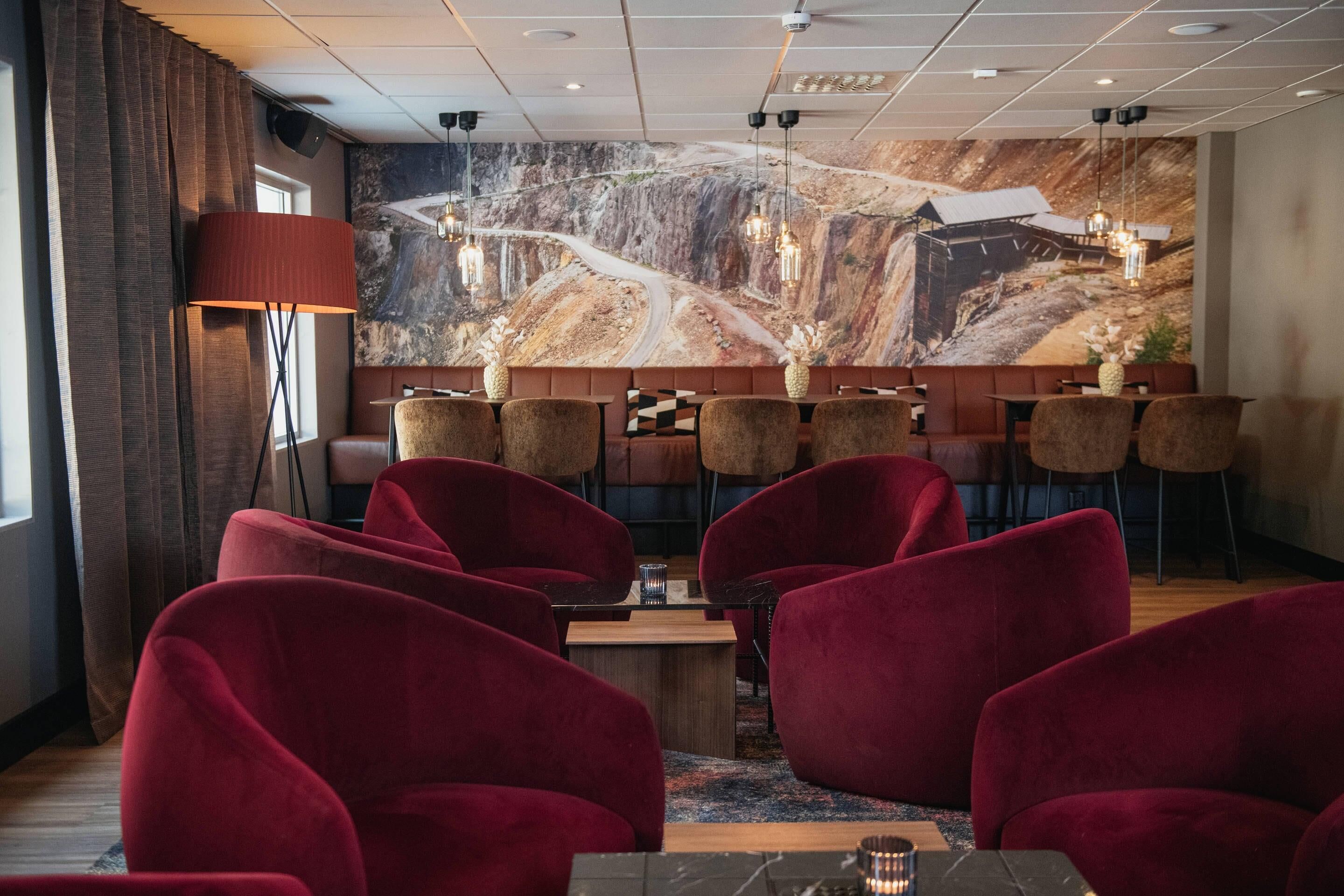 Restaurante com cadeiras estofadas vermelhas, um mural grande de uma estrada de montanha e iluminação pendente.