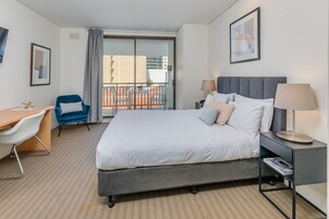 Quarto de hotel com cama, escrivaninha, cadeira, mesa de cabeceira e varanda com vista para edifícios.