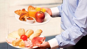 Uma pessoa segurando um prato com croissants, um muffin, um copo de suco de laranja, uma maçã e outro prato com queijo, presunto e pãezinhos.
