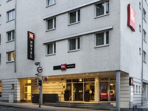 Um hotel com fachada moderna, um destaque para a placa 'ibis' e uma entrada visível com portas de vidro.